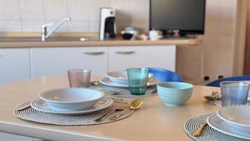 Uma mesa de jantar bem posta com pratos, tigelas e copos, uma cafeteira e uma televisão ao fundo.