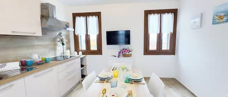 Cozinha compacta com armários brancos, fogão embutido e espaço de jantar para quatro pessoas, com mesa, cadeiras e televisão fixada na parede.