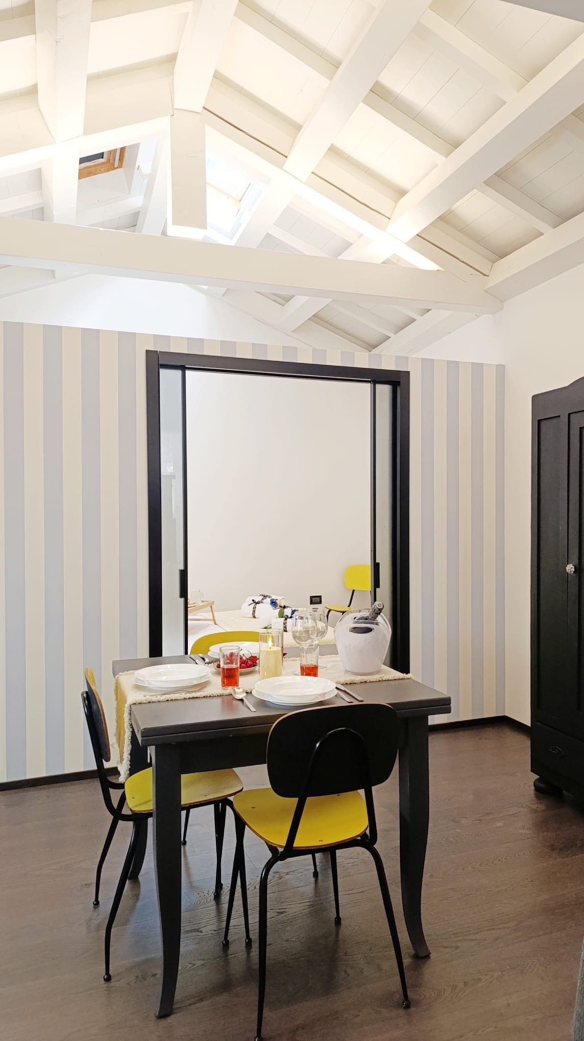 Uma sala de jantar com mesa posta para quatro pessoas, papel de parede listrado e uma porta preta.