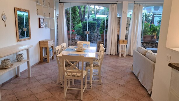 Uma sala de jantar com mesa e cadeiras, um espelho e vista para o jardim através de amplas janelas.