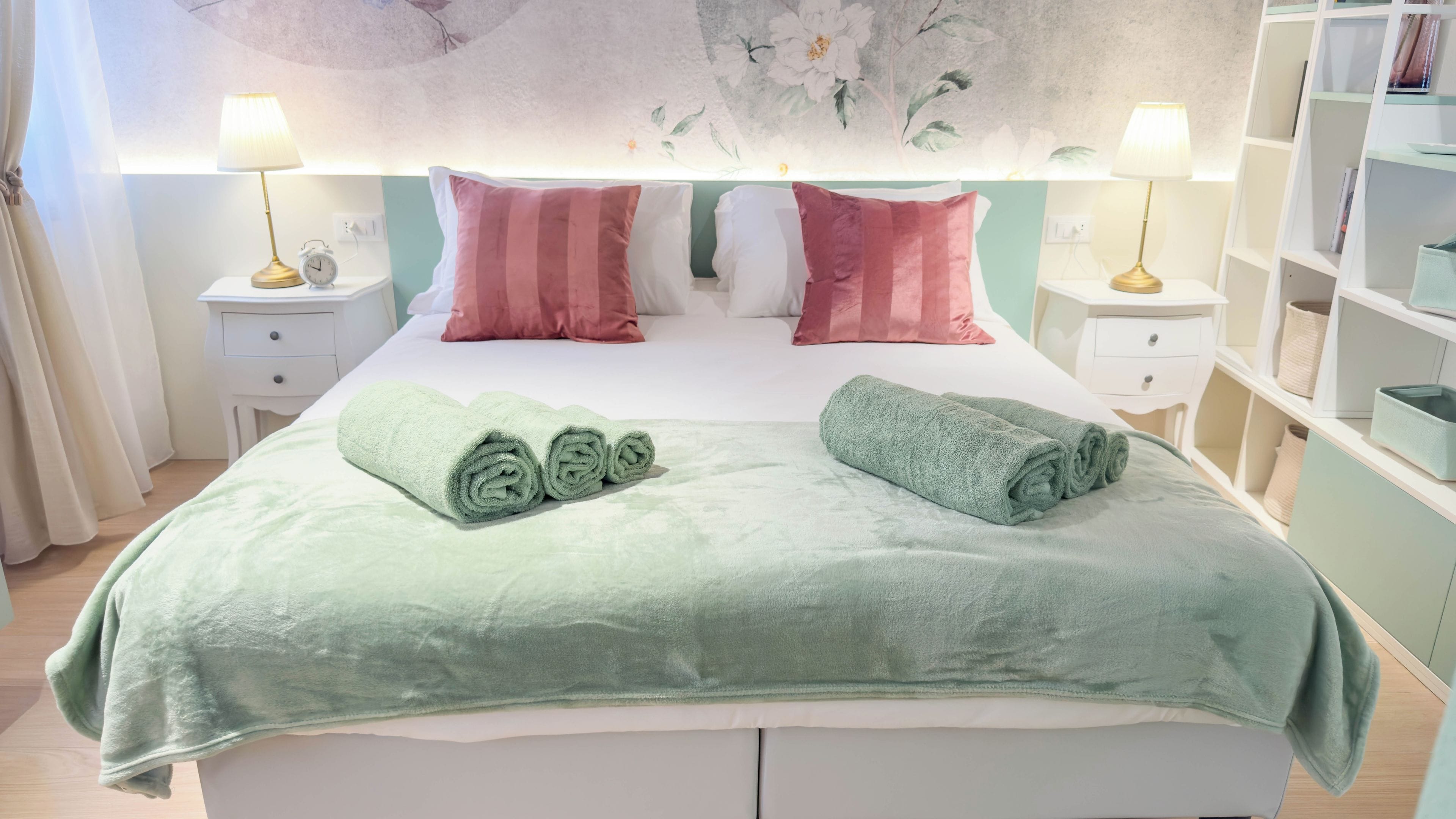 Cama bem arrumada com toalhas enroladas, cabeceira branca e um mural floral na parede.