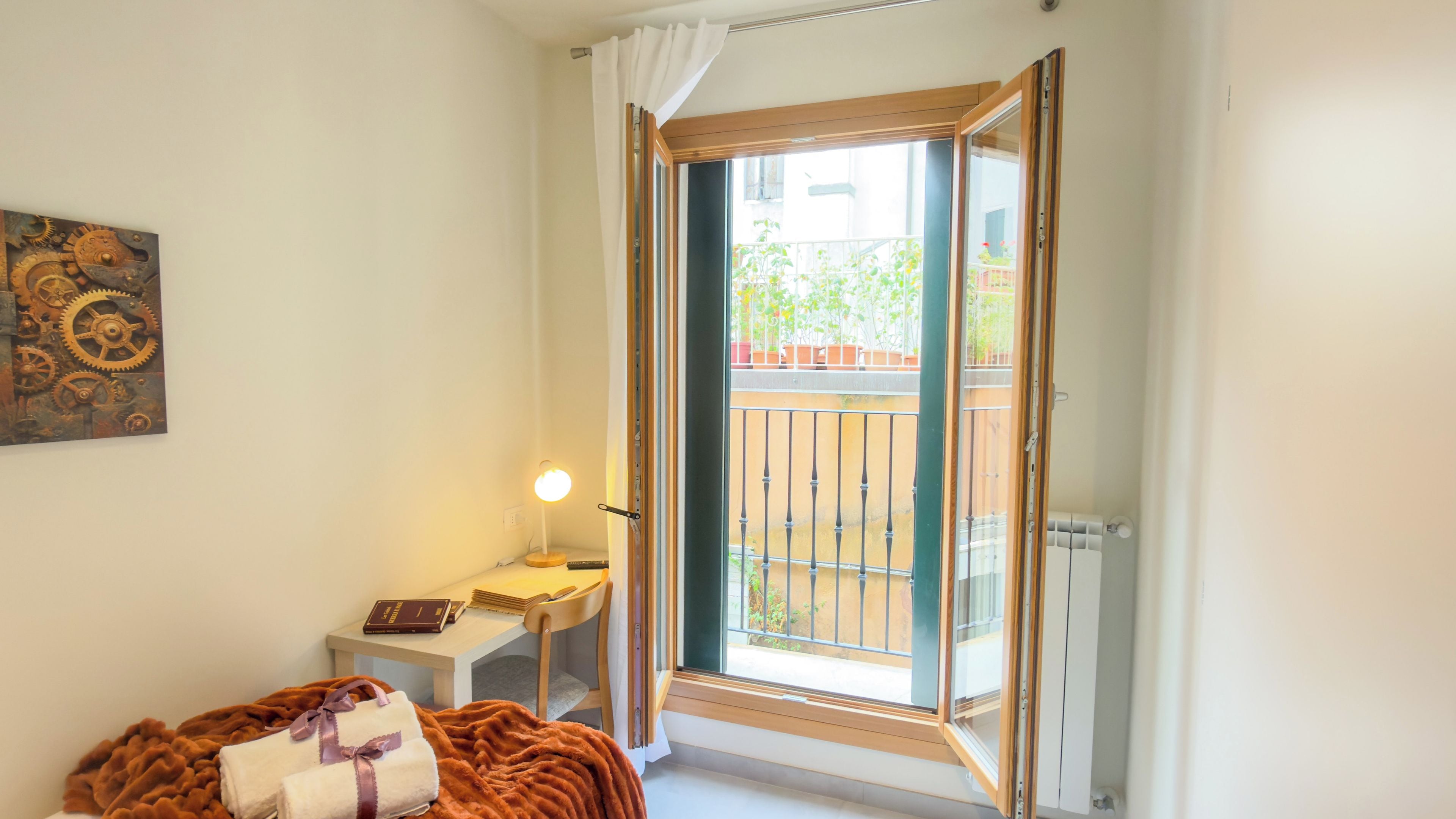 Quarto com cama, mesa de cabeceira com luminária, radiador e janela com vista para uma varanda.