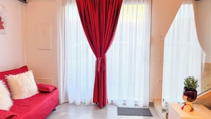 Quarto com sofá vermelho, cortinas brancas e uma pequena planta em uma mesinha lateral.