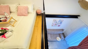 Quarto com cama, mesa de cabeceira e um banquinho pequeno. Há uma pintura na parede e uma janela com cortinas.