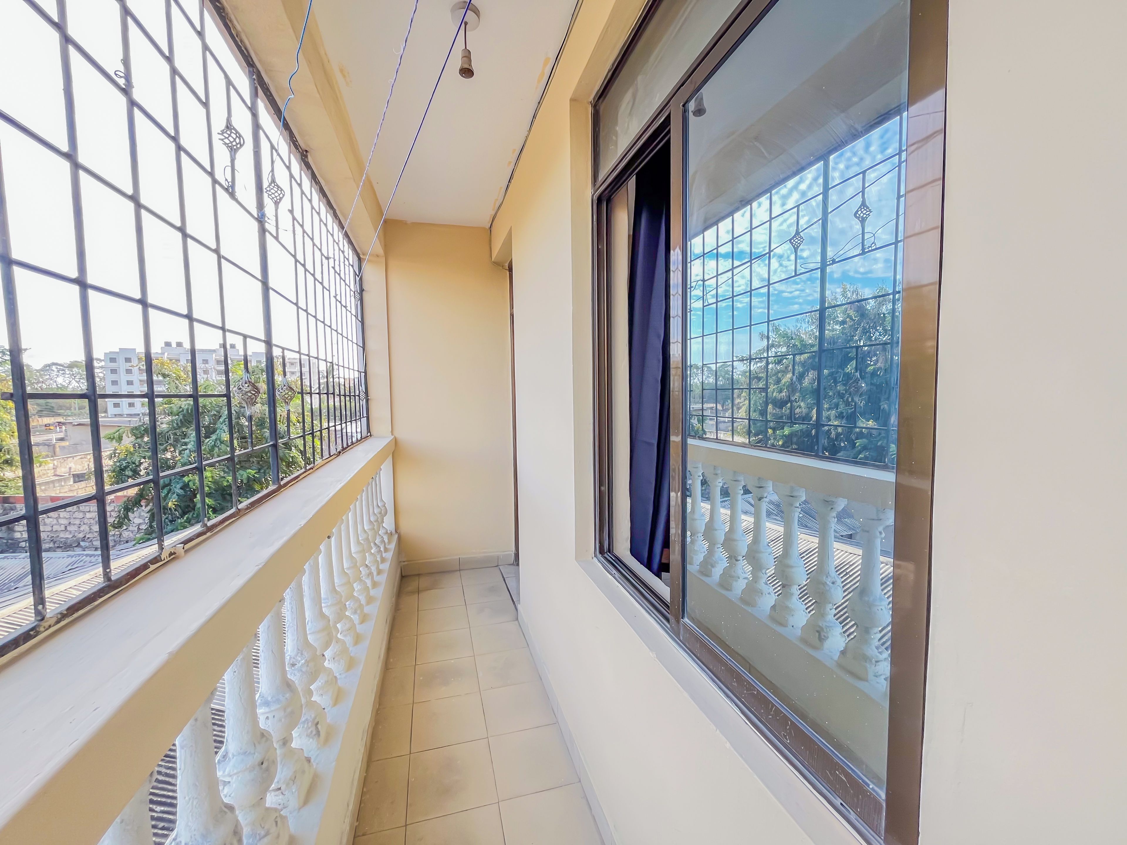 Um corredor estreito com piso de azulejos, uma varanda com grade de metal e uma janela com cortina.