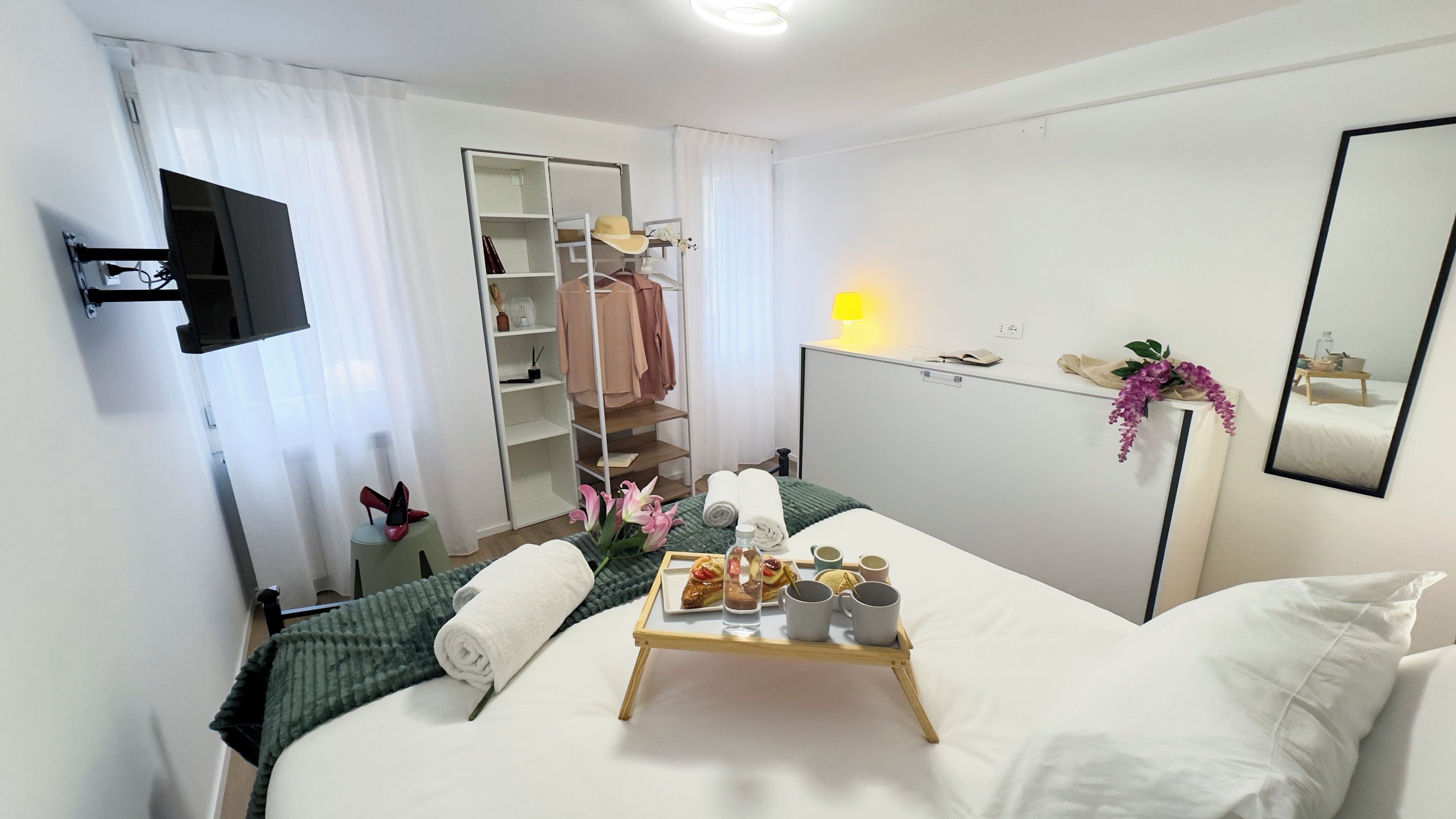 Quarto de hotel com cama, mesa de cabeceira com bandeja de comida e bebidas, armário, espelho e televisão.