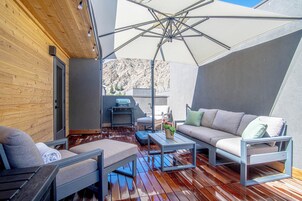 Uma área de estar moderna ao ar livre, com deck de madeira, um grande guarda-sol e vista para uma paisagem montanhosa.