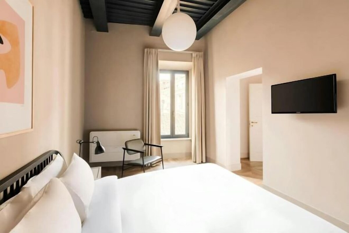 Quarto de hotel com cama, poltrona, televisão e janela com cortinas.