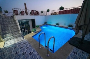Piscina no terraço com estrutura de vidro, rodeada por plantas em vasos e móveis de exterior.