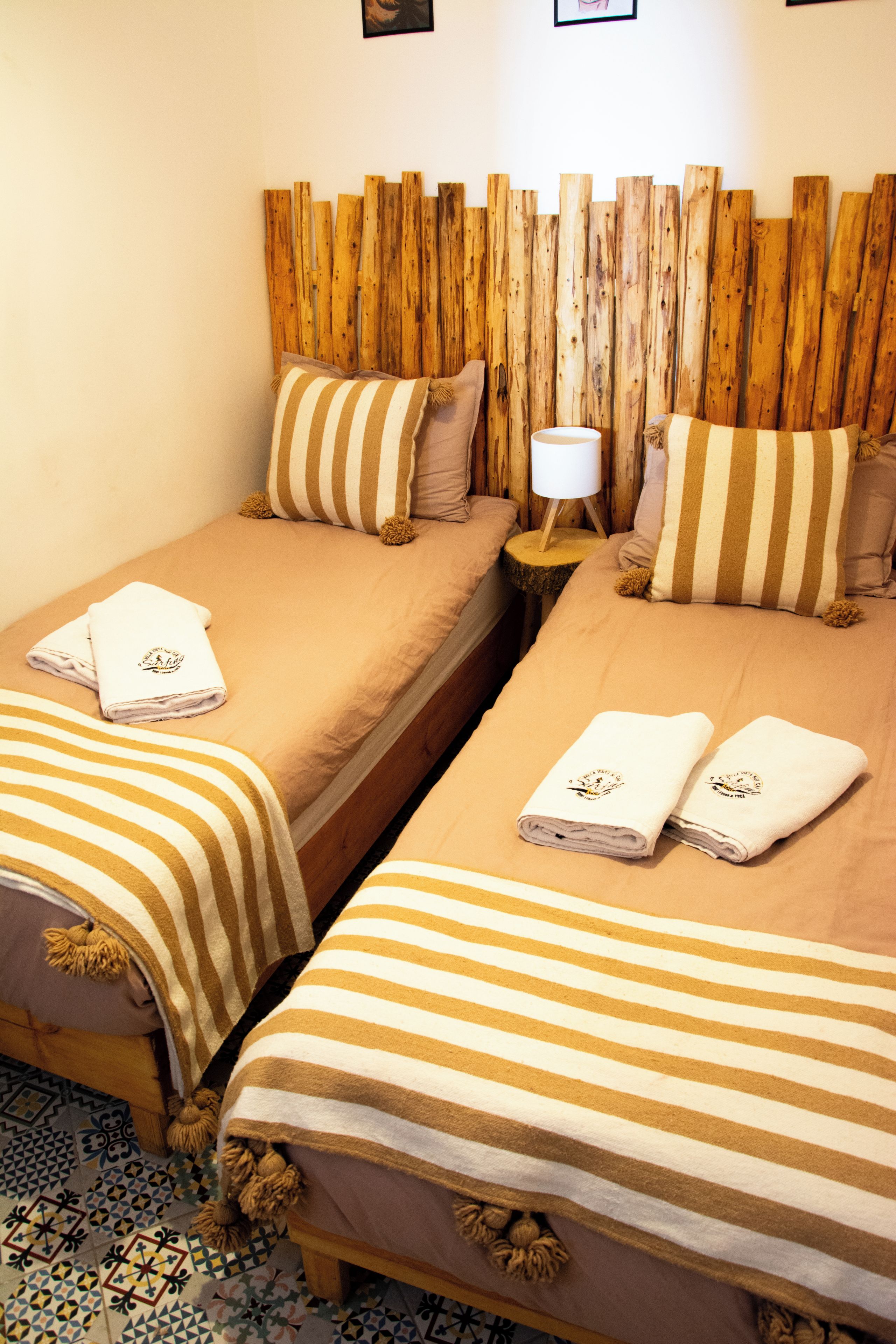 Quarto de hotel com duas camas, cada uma com roupa de cama e almofadas listradas, mesa de cabeceira com abajur e cabeceira de madeira.