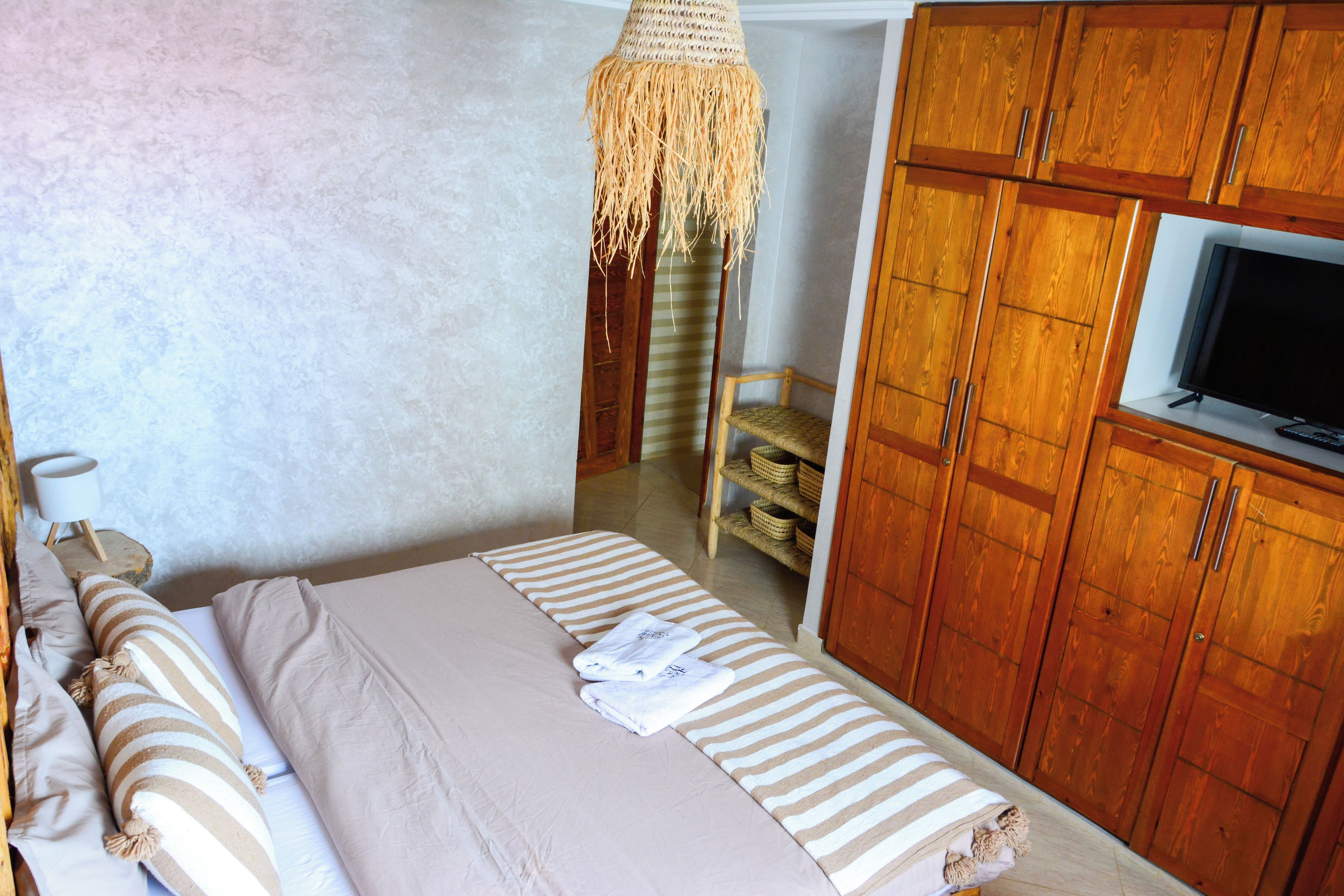 Quarto com cama, televisão sobre um armário de madeira e luminária pendente de fibra.