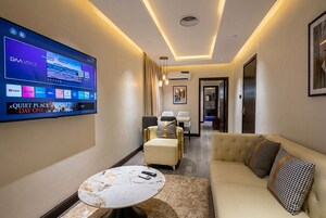 Quarto de hotel com televisão de tela plana exibindo um serviço de streaming, um sofá, uma mesa de centro redonda e um corredor que leva a uma área de refeições.
