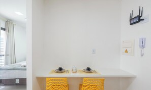 Quarto de hotel com cama, escrivaninha e duas cadeiras, telefone fixado na parede e tomada.
