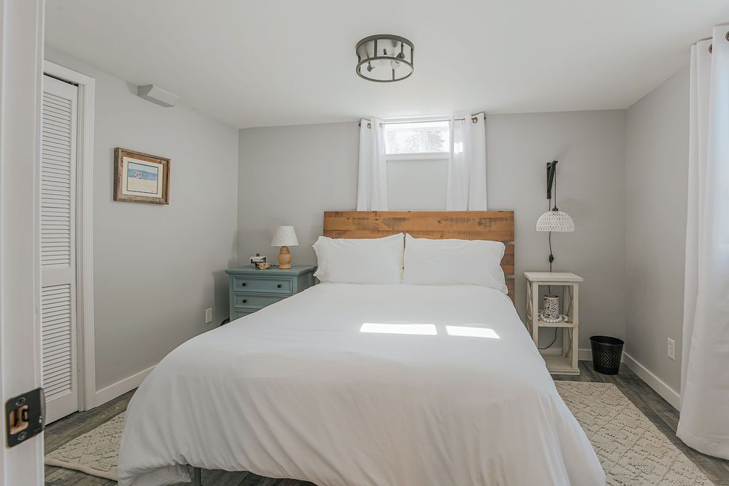 Quarto com cama, mesa de cabeceira, luminária, quadro na parede e janela com cortinas.