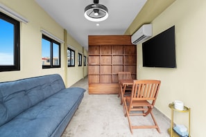 Quarto com sofá azul, estante de madeira, mesa com cadeiras e televisão na parede.