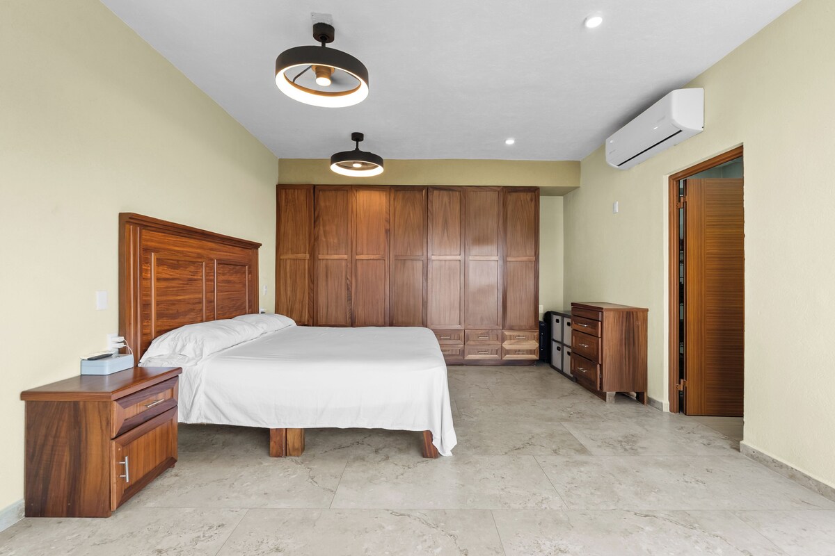 Quarto com cama, guarda-roupa de madeira, mesas de cabeceira e luminária no teto.