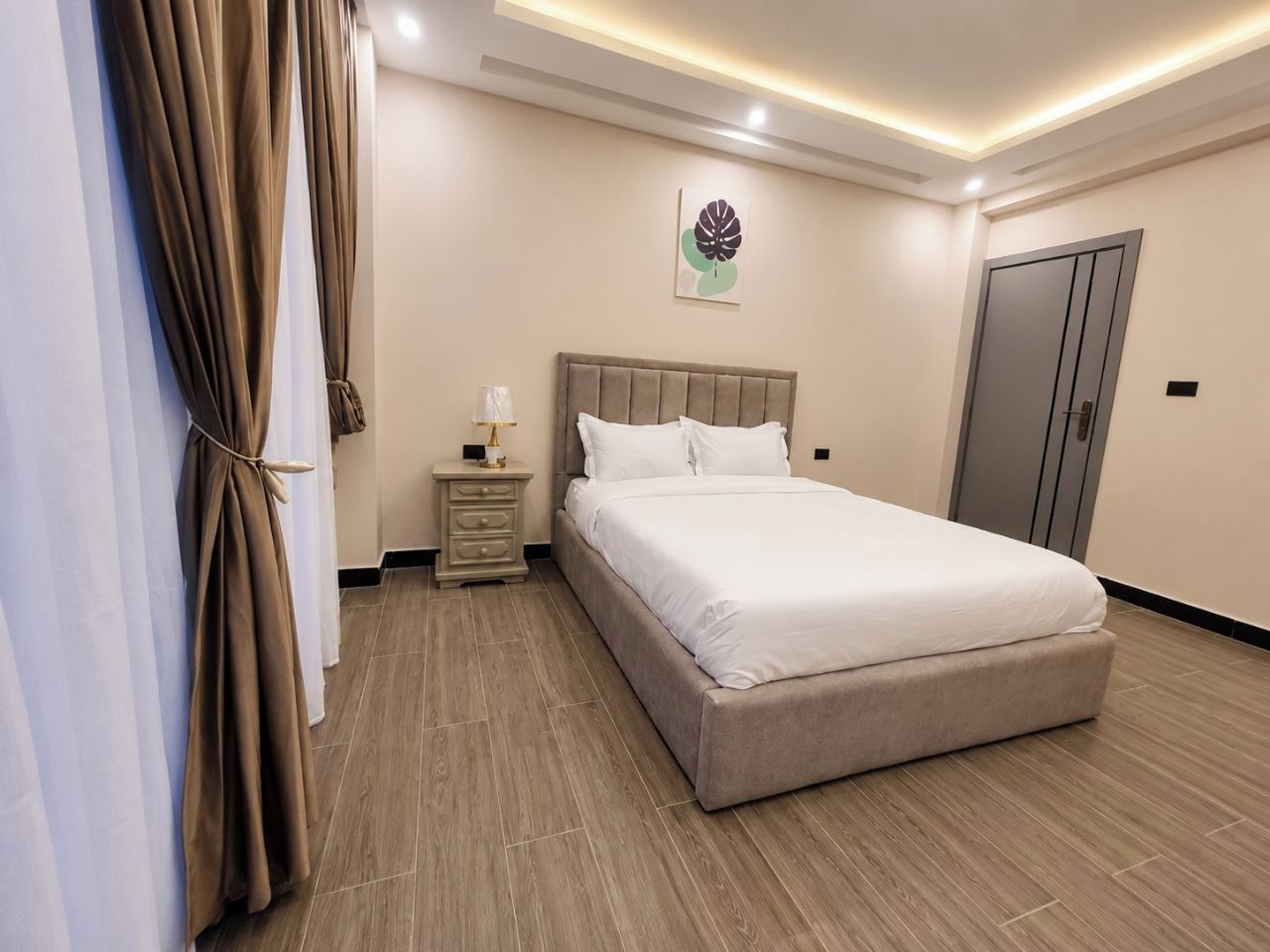 Quarto com cama, mesa de cabeceira, cortinas e uma obra de arte na parede.