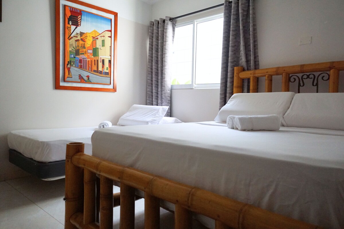 Quarto de hotel com uma cama grande, uma cama menor, cabeceira de madeira, janela com cortinas e um quadro na parede.