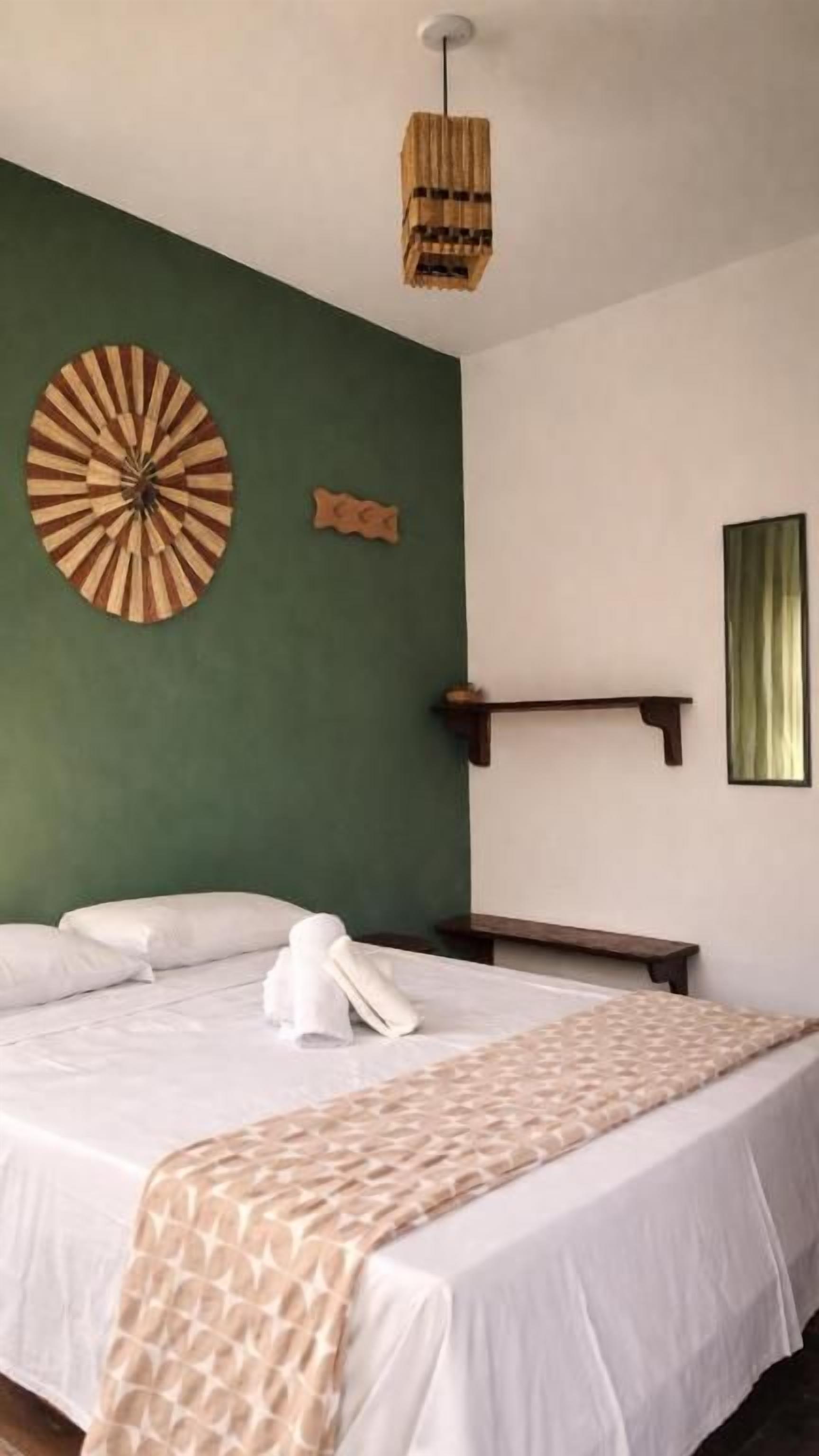 Cama bem arrumada com edredom branco e manta decorativa, um relógio de parede e luminária pendente.