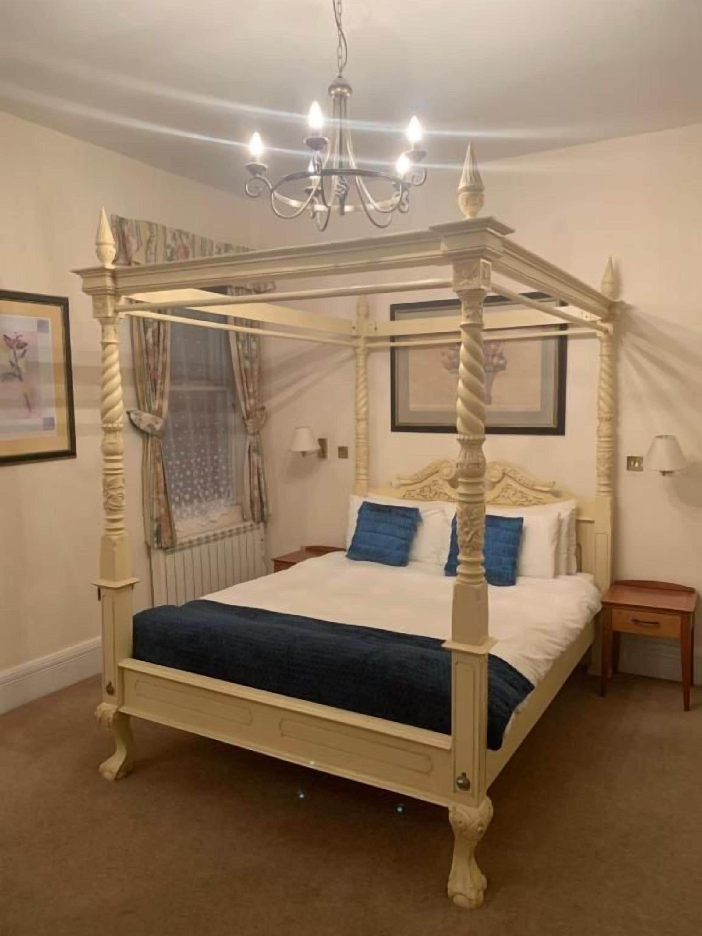 Uma cama com dossel e colunas, estrutura branca e roupa de cama azul, uma mesa de cabeceira e uma luminária fixada na parede.