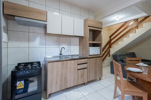 Cozinha moderna com fogão a gás, forno e micro-ondas embutido. Há uma mesa de jantar de madeira com cadeiras e um sofá na área de estar adjacente.