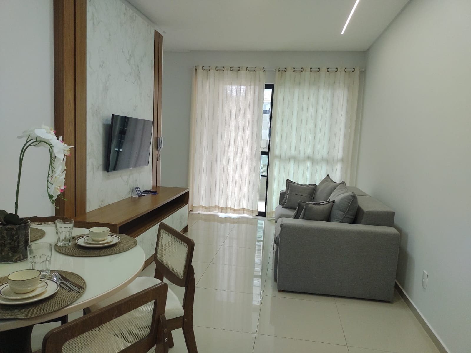 Sala de estar moderna com um sofá cinzento, área de jantar com mesa posta para dois e TV na parede.