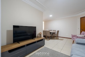 Uma sala moderna com televisão de tela plana em um móvel de madeira, área de jantar com mesa e cadeiras, e quarto com cama e uma cadeira rosa.