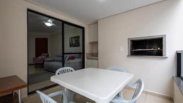 Uma área de refeições com uma mesa branca e cadeiras azuis, uma porta de vidro deslizante que dá para um quarto, e uma pequena cozinha com adega embutida.