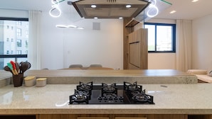 Uma cozinha moderna com bancada de granito, fogão a gás e forno embutido. Há uma janela com cortinas, um armário de madeira e uma área de jantar com mesa e cadeiras.