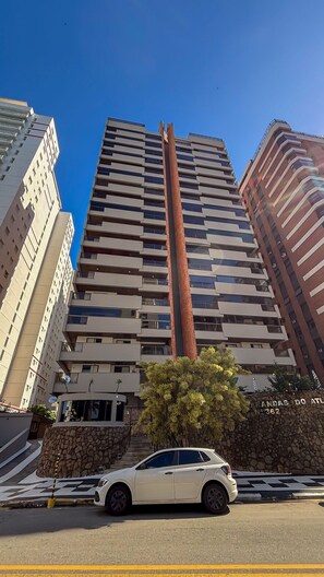 Um edifício alto com um elemento vertical central, um carro branco estacionado na frente e um muro de pedra com uma placa.