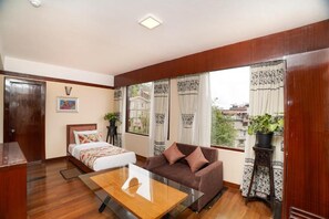 Quarto de hotel com cama, sofá, mesa de centro e grandes janelas com cortinas.
