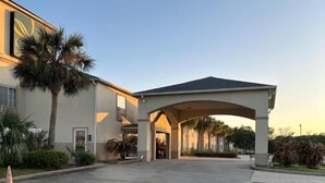 Entrada de hotel com acesso coberto, palmeiras e céu claro.