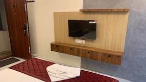 Quarto de hotel com cama, televisão fixada num painel de madeira, porta e pequena estante acima da TV.