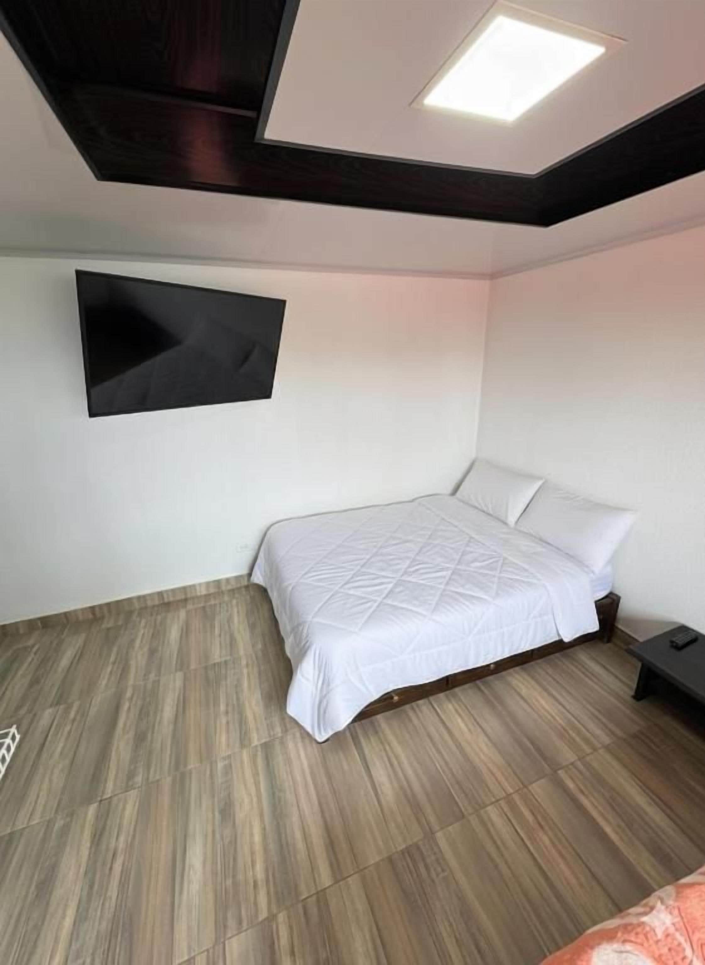 Quarto com cama, TV fixada na parede e piso de madeira.