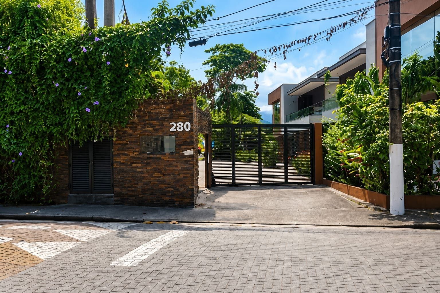 Uma entrada fechada por portão, com muro de tijolos e placa com o número '280', conduz a um edifício moderno com janelas de vidro e portão de metal.