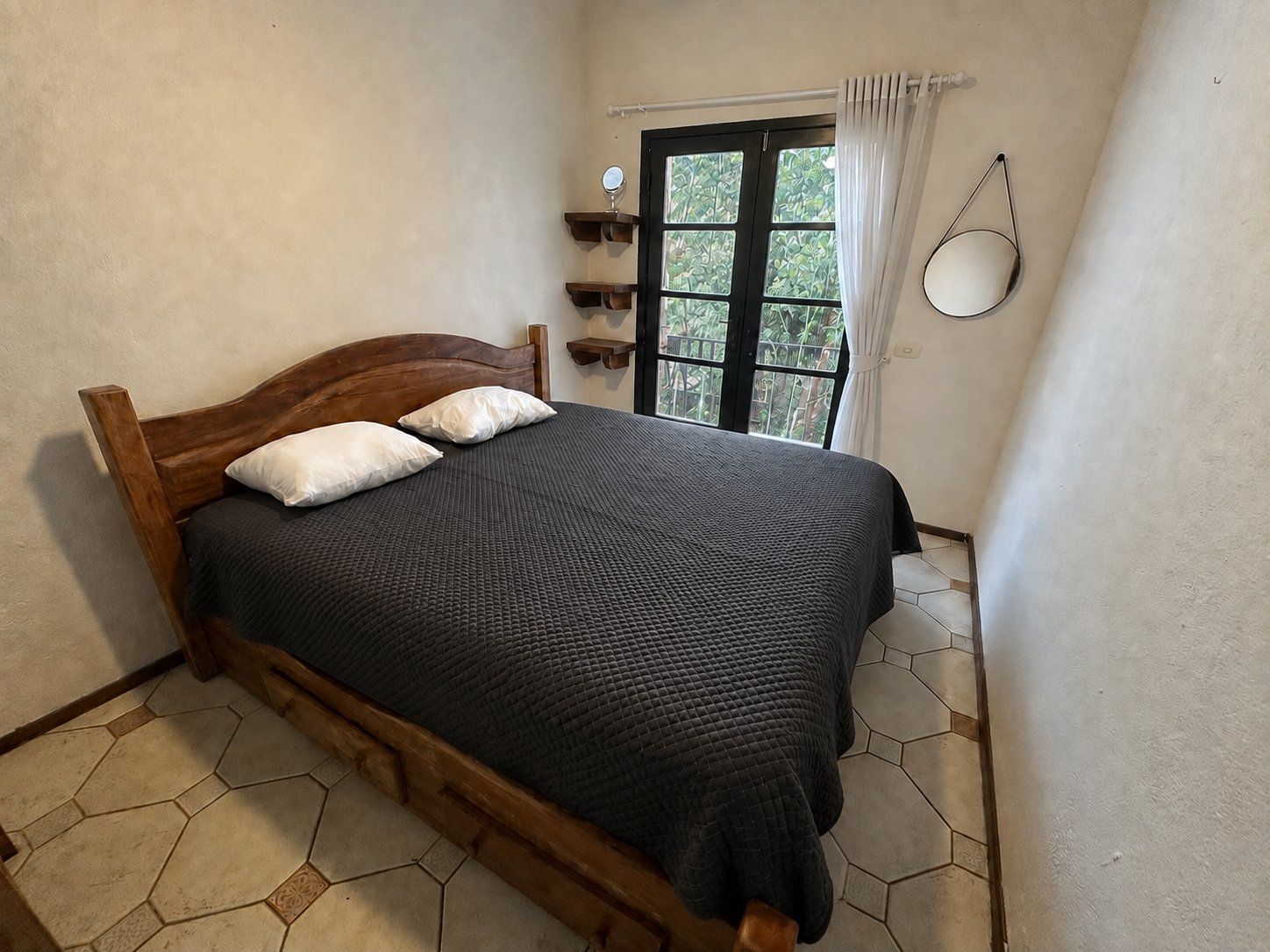 Quarto com cama, janela com cortinas, espelho e vista para o verde lá fora.