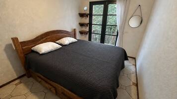 Quarto com cama, janela com cortinas, espelho e vista para o verde lá fora.