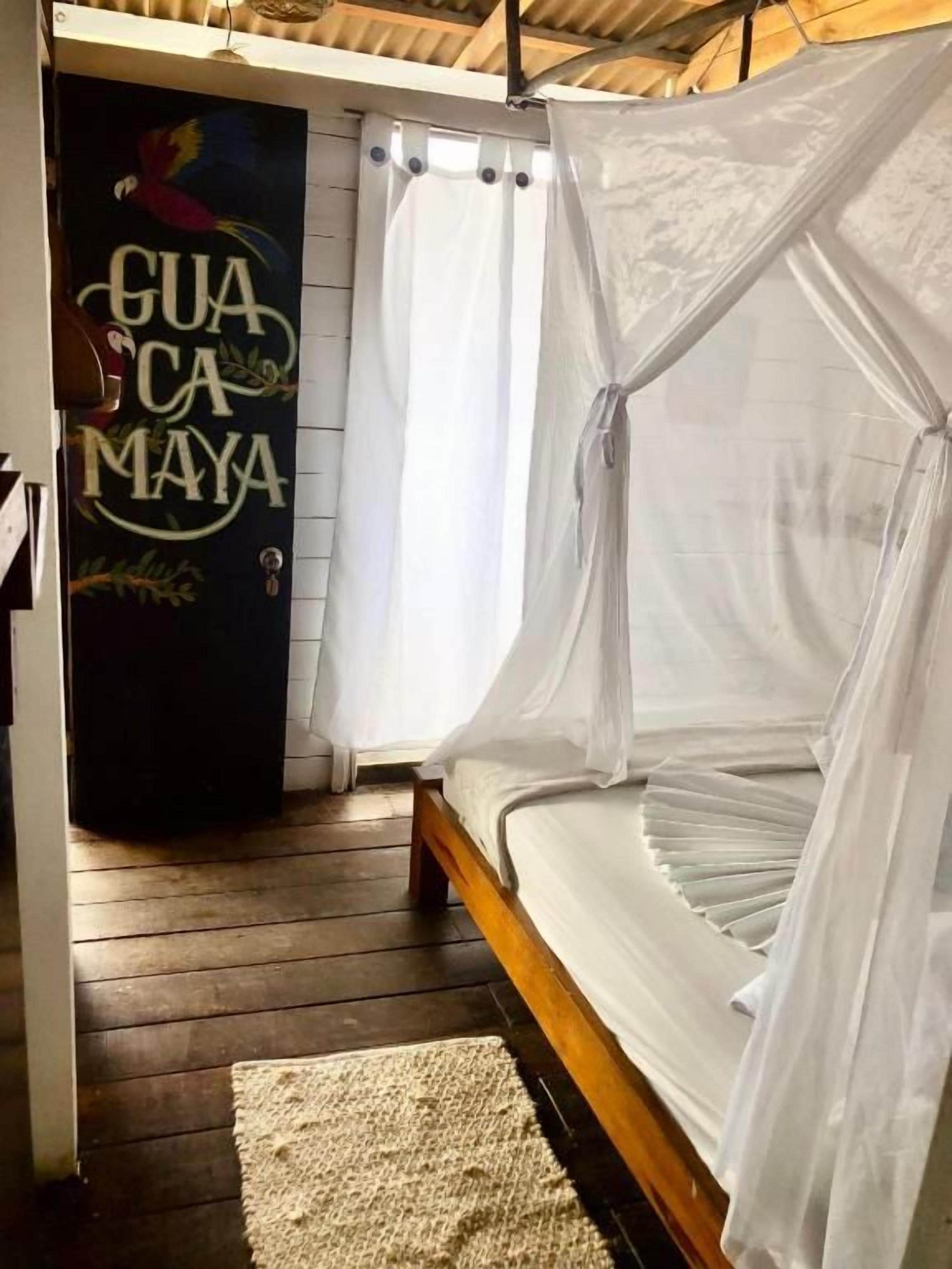 Quarto com cama coberta por mosquiteiro, piso de madeira e parede com painel decorativo.