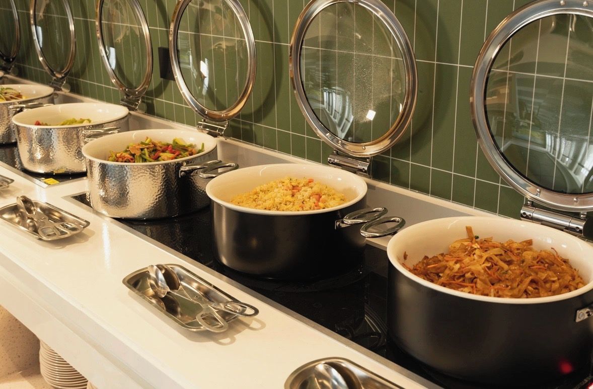 Buffet com diversos pratos em recipientes de metal e cerâmica, bancada branca e parede com azulejos verdes.