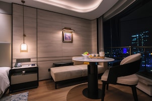 Quarto de hotel moderno com cama, escrivaninha, cadeira e mesa redonda com uma tigela de frutas.