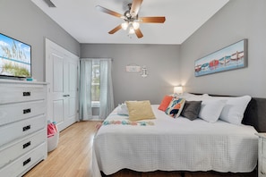 Um quarto com cama, ventilador de teto, televisão, quadro e porta.