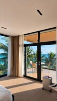 Quarto de hotel moderno com uma grande porta de vidro deslizante que dá para uma varanda com vista para palmeiras e o oceano.