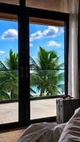 Quarto com grande porta de correr de vidro que dá para uma varanda com vista para palmeiras e praia.