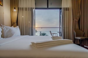 Quarto de hotel com cama, uma cadeira, mesa pequena e vista para o mar através de uma grande janela.