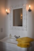 Casa de banho com lavatório branco, espelho com moldura ornamentada, duas luzes na parede e toalha amarela sobre o lavatório.