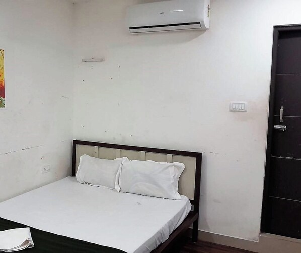 Uma cama de solteiro com colchão e travesseiros brancos, ar-condicionado fixado na parede e uma porta fechada.