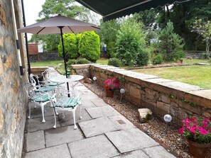 Um pátio externo com piso de pedra, uma mesa e cadeiras, uma pérgola com cobertura e um muro de pedra com vasos de plantas.