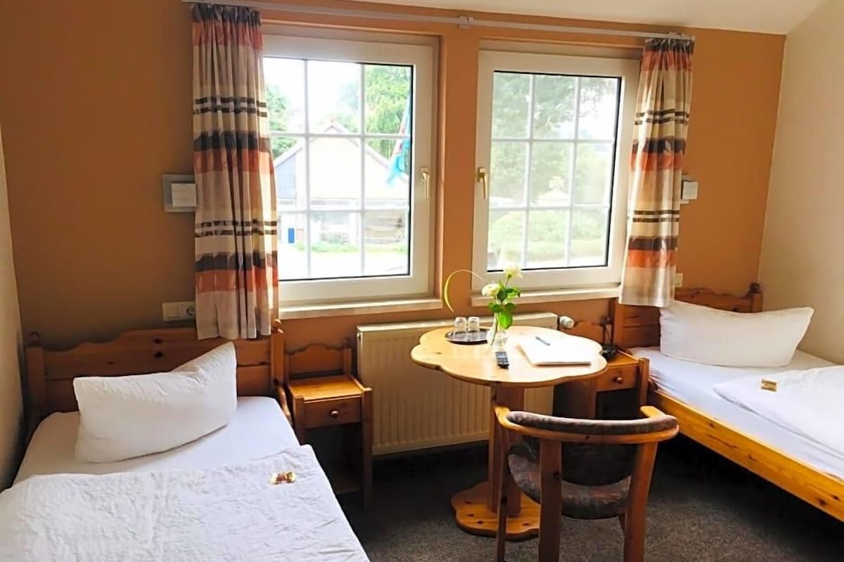 Quarto de hotel com duas camas, uma mesinha, uma cadeira e uma janela com cortinas.