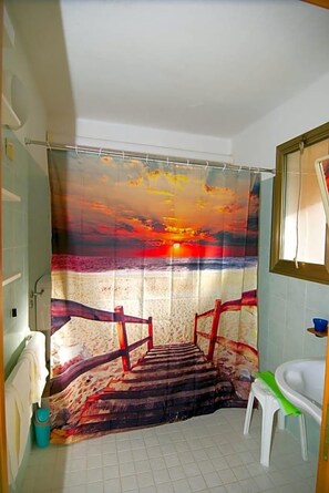 Casa de banho com cortina de chuveiro retratando um pôr do sol sobre a praia, uma pia branca e um banquinho verde.