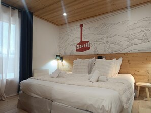 Quarto com cama grande, cabeceira com painel de madeira, mural de montanhas na parede, um lampião vermelho e um banquinho pequeno.